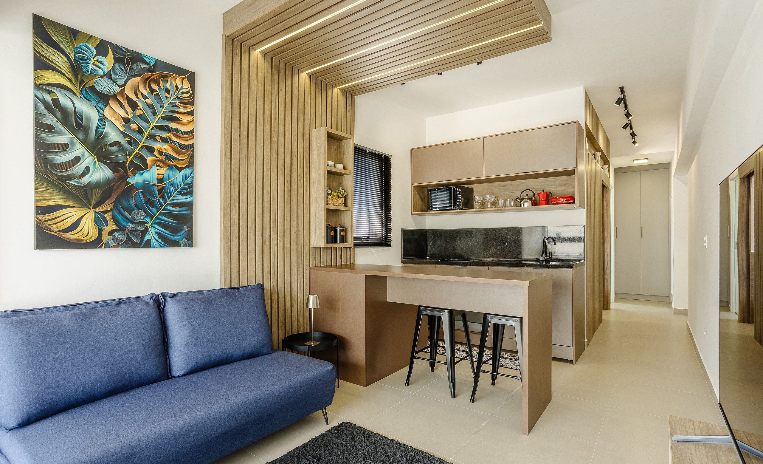 Uma sala de estar moderna com um sofá azul, uma cozinha com espaço para bar e uma grande obra de arte com folhagem tropical na parede.