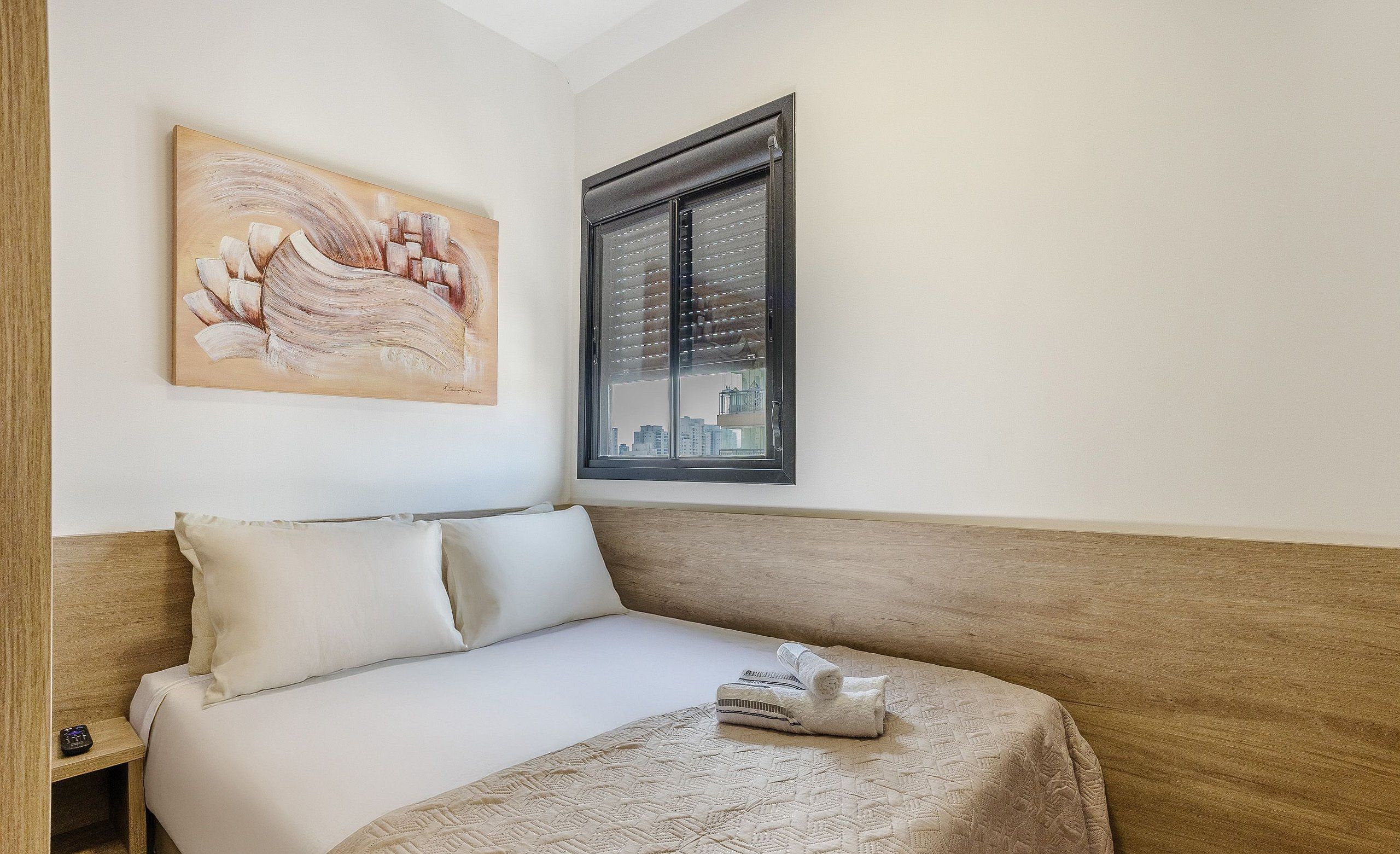 Cama bem arrumada com um cobre-leito bege, um travesseiro e uma toalha dobrada em uma cabeceira de madeira. Um quadro está pendurado na parede acima da cama.