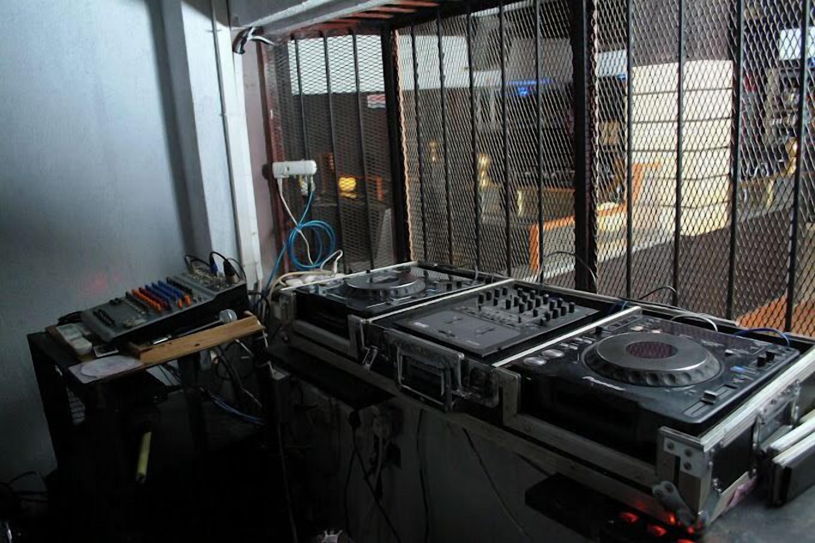 Equipamento de DJ com mixer, toca-discos e cabos, posicionado perto de uma janela com tela de proteção metálica.