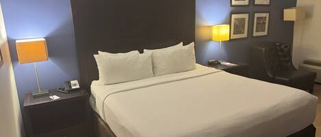 Quarto de hotel com uma cama grande, mesa de cabeceira com abajur, uma cadeira e quadros nas paredes.