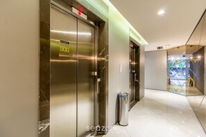 Elevador moderno com placa indicando acessibilidade, uma lixeira e um corredor que leva a uma área com paredes de vidro.