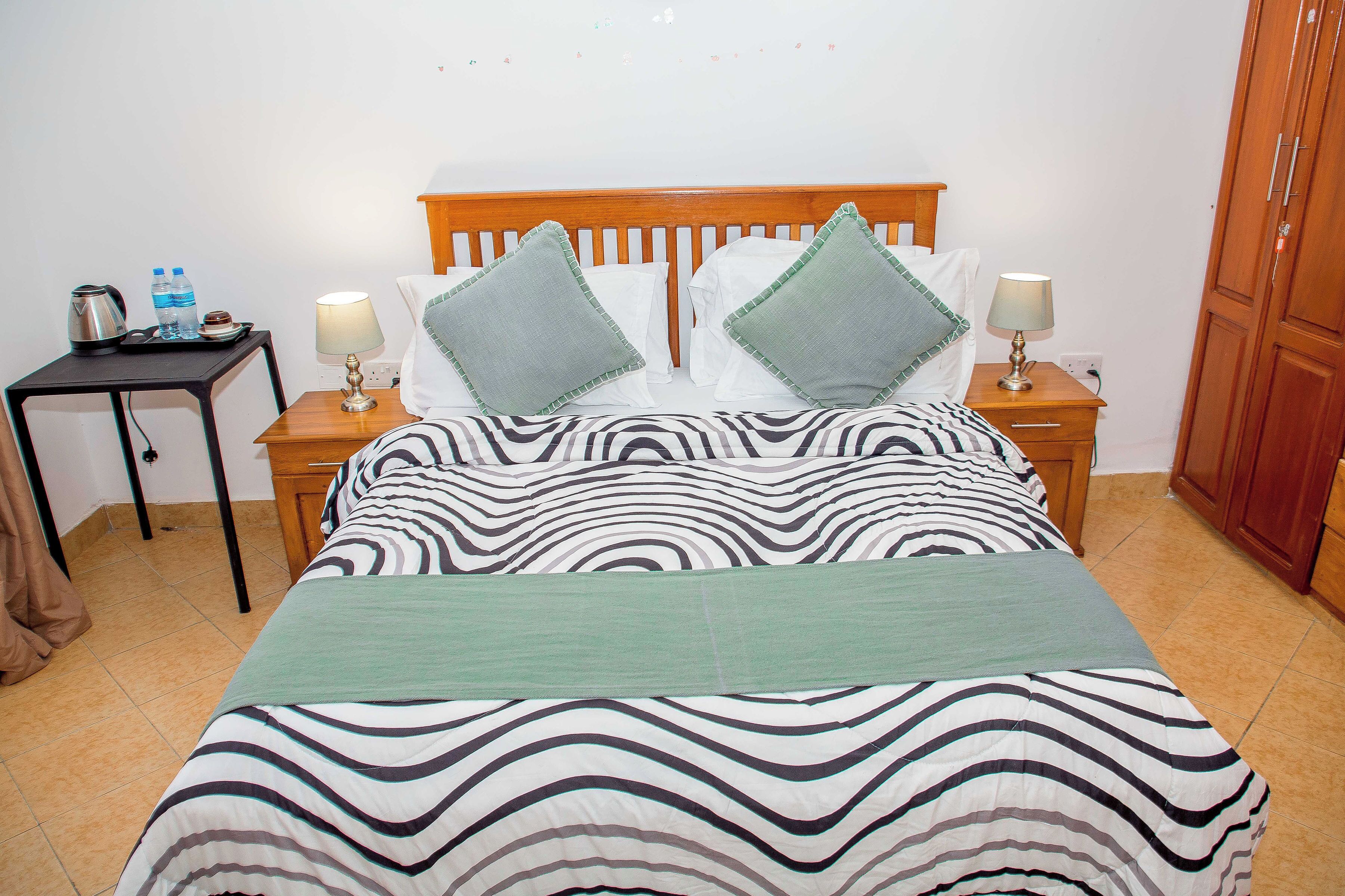 Quarto de hotel com cama, duas mesas de cabeceira, um criado-mudo, um guarda-roupa de madeira e chão ladrilhado.