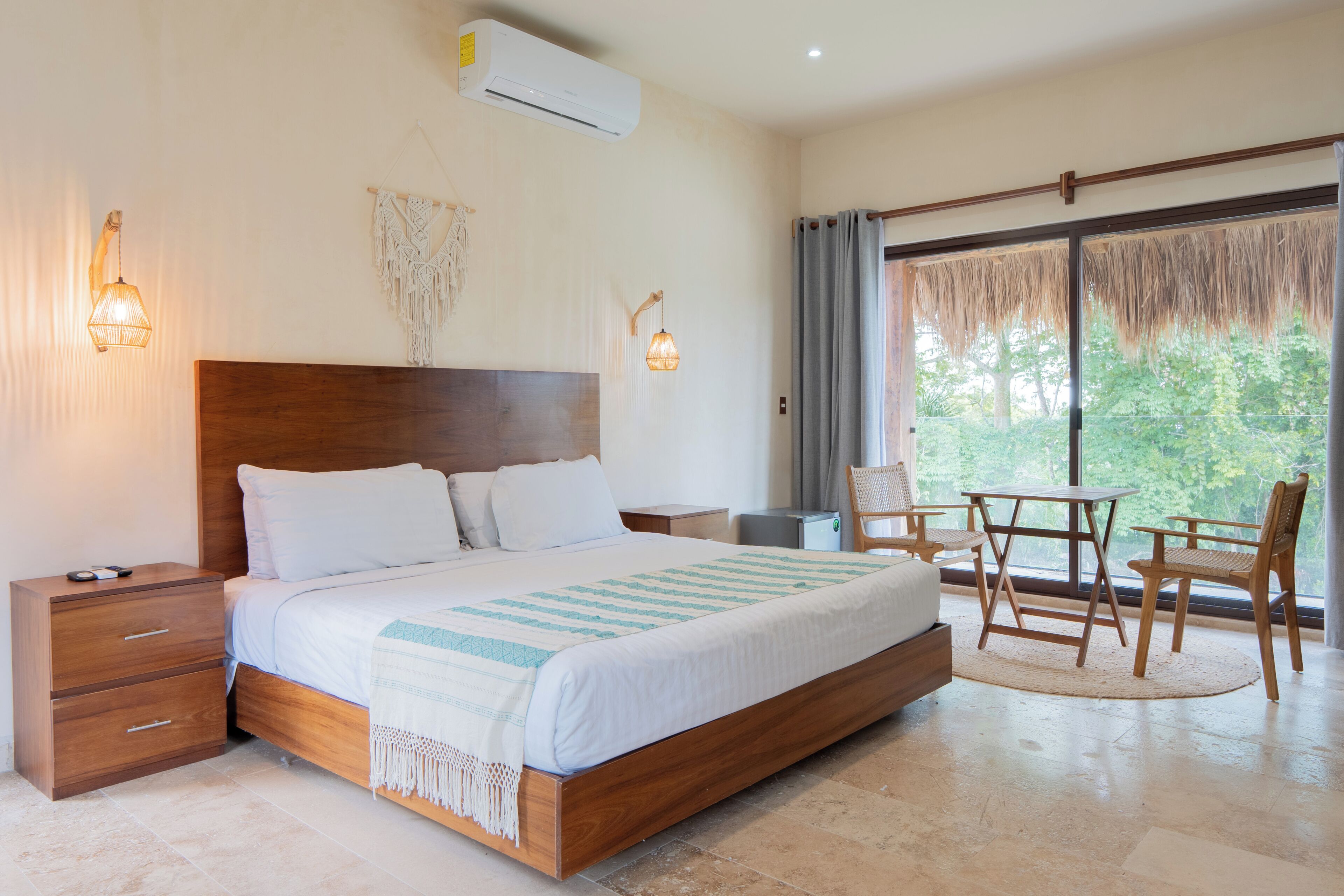 Quarto de hotel com uma cama grande, mesas de cabeceira, escrivaninha, cadeira, janela com cortinas e ar condicionado.