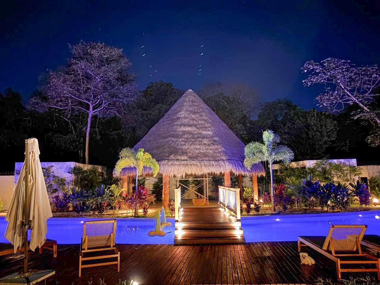 Estrutura com telhado de palha, piscina e deck de madeira, cercada por plantas tropicais.