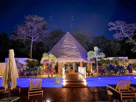 Estrutura com telhado de palha, piscina e deck de madeira, cercada por plantas tropicais.