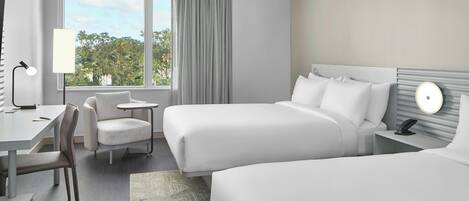 Quarto de hotel com duas camas, uma mesa, uma cadeira, um abajur e vista para árvores pela janela.