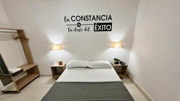 Quarto de hotel moderno com cama, abajures, uma mesinha com uma planta e um letreiro fixado na parede.