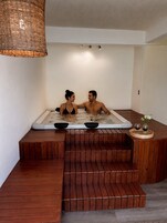 Uma banheira de hidromassagem com degraus de madeira que a levam, duas pessoas sentadas dentro, e um tabuleiro com copos ao lado.