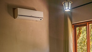 Uma cama bem arrumada com cabeceira de madeira, um aparelho de ar condicionado branco na parede e uma janela com cortina.