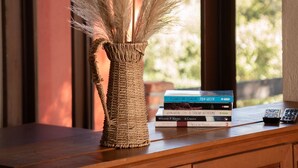 Uma mesa de madeira com um vaso trançado contendo plumas de capim-do-campo, uma pilha de livros e um controle remoto.