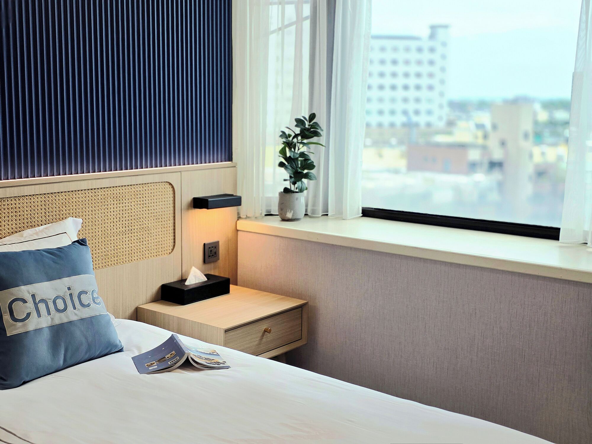 Quarto de hotel com cama, mesa de cabeceira com abajur, janela com vista para edifícios e uma planta no parapeito.