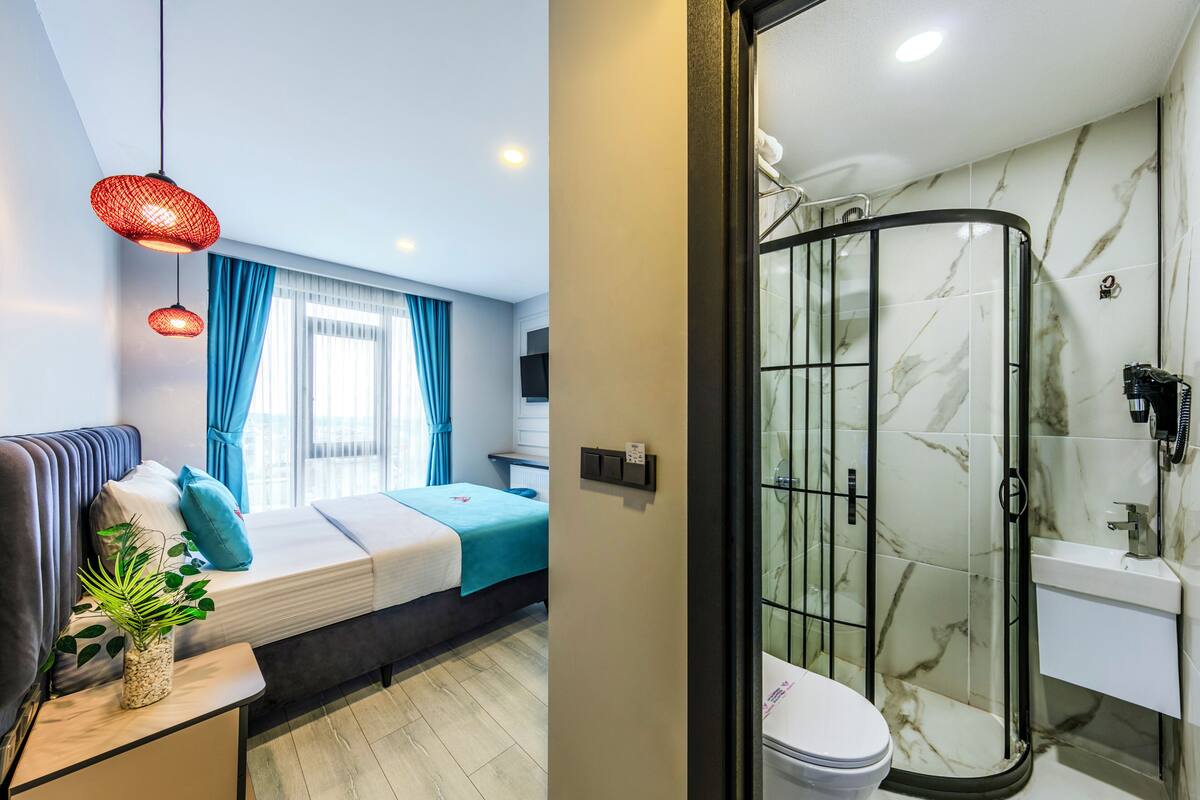 Quarto moderno com cama, mesa de cabeceira e planta em vaso. Inclui banheiro com box de vidro e vaso sanitário.