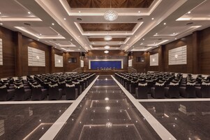 Uma sala de conferências com fileiras de cadeiras, palco e um lustre.