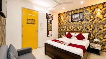 Quarto de hotel com cama, sofá, tapete de boas-vindas, ventoinha no teto e parede com padrão de folhas.