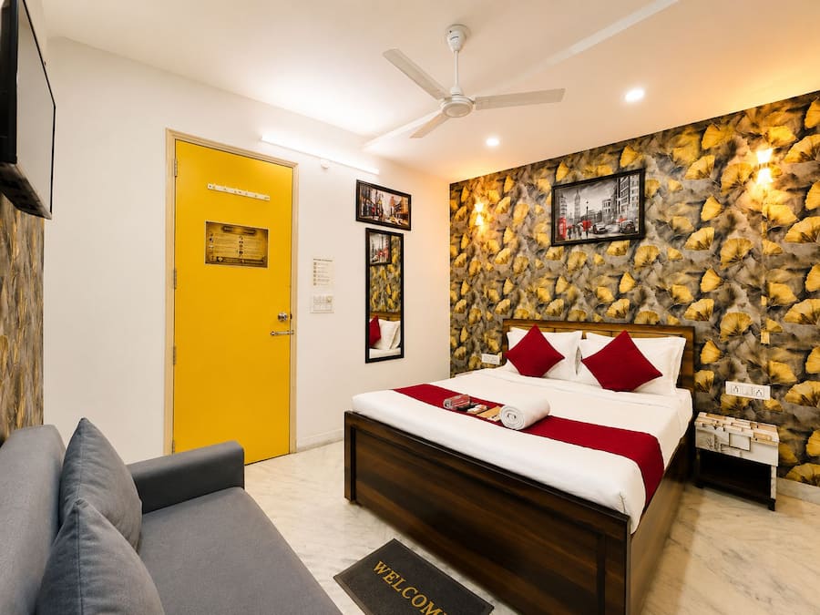 Quarto de hotel com cama, sofá, tapete de boas-vindas, ventoinha no teto e parede com padrão de folhas.