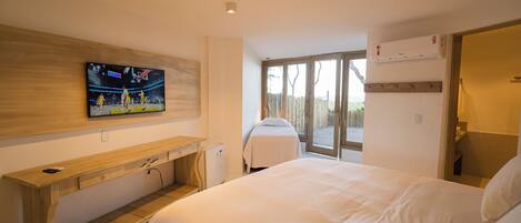 Quarto de hotel com cama, televisão fixada na parede, secretária e janela com vista para o exterior.