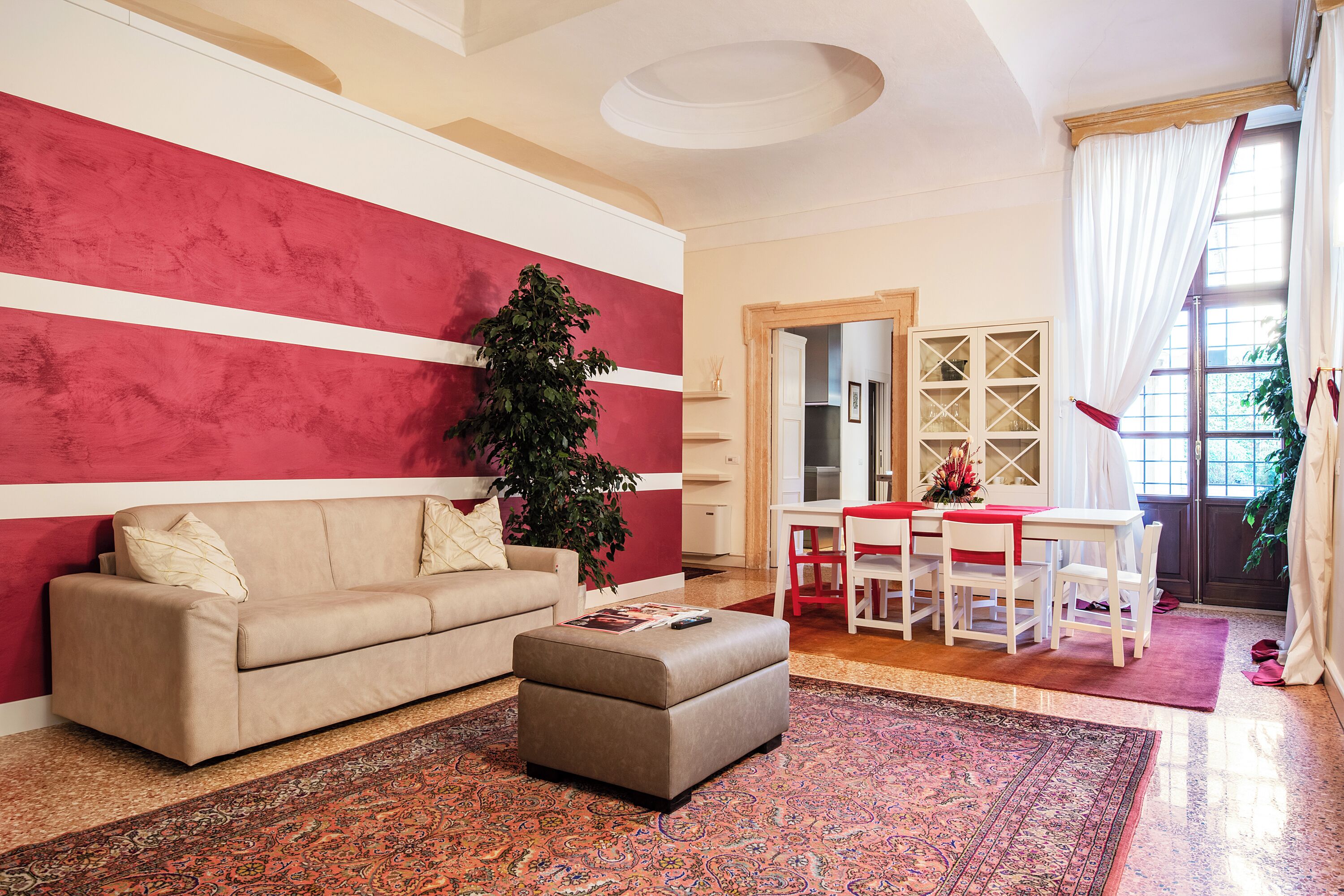 Uma sala de estar com sofá bege, parede em destaque vermelha, planta grande, área de jantar com mesa e cadeiras brancas, e tapete estampado.