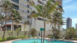 Área de piscina com palmeiras, espreguiçadeiras e um edifício com varandas ao fundo.