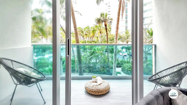 Varanda com vista para palmeiras e um pequeno vaso de planta. Há duas cadeiras pretas e um sofá cinza.