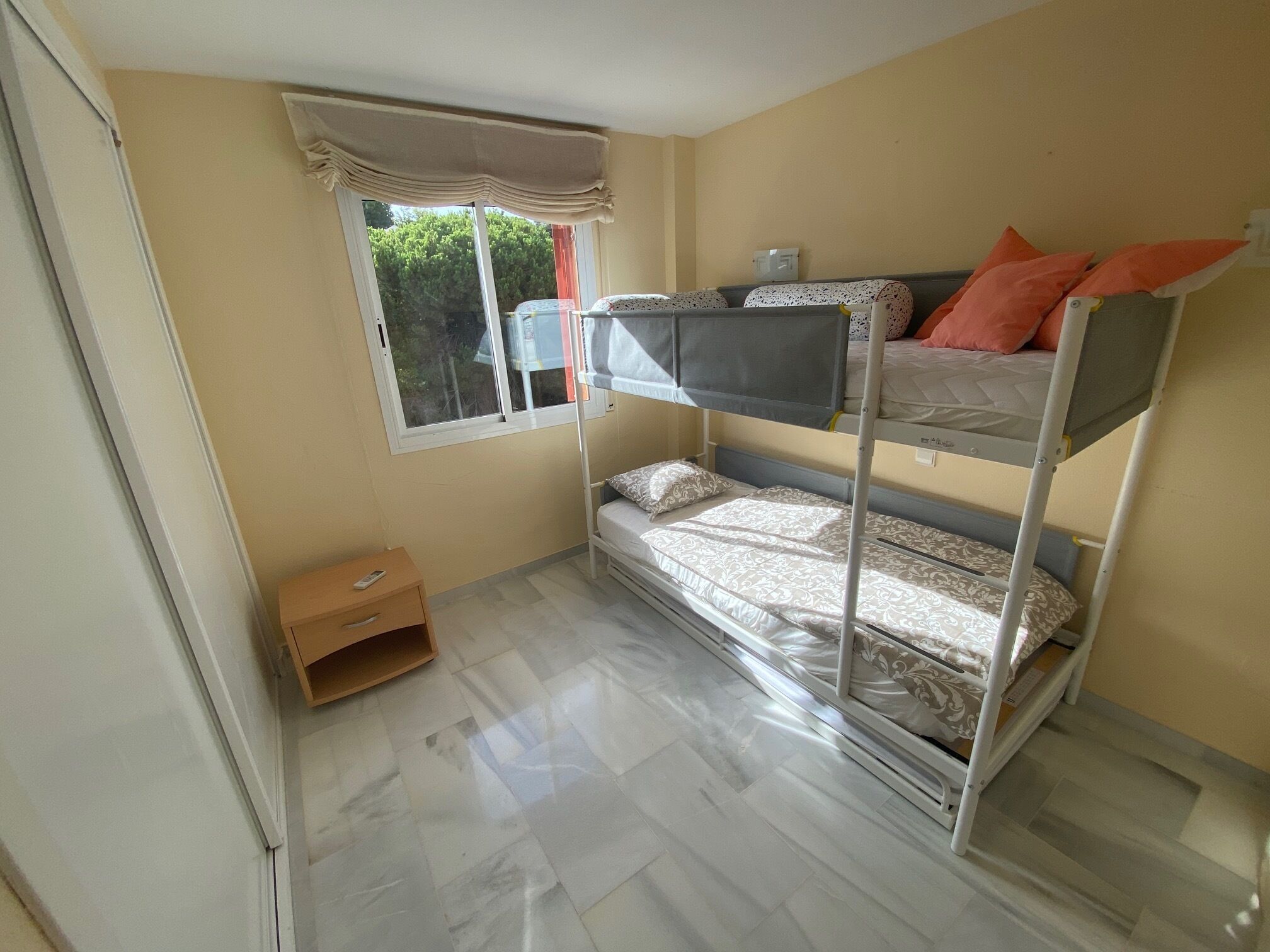 Quarto pequeno e bem iluminado com duas beliches, uma janela com cortinas, uma pequena mesa de madeira e piso ladrilhado.