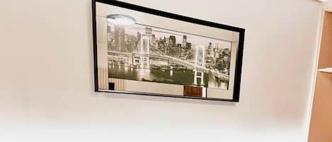 Quarto de hotel com uma cama grande, uma cama menor, uma mesinha de cabeceira com uma lâmpada e um quadro com uma vista urbana na parede.