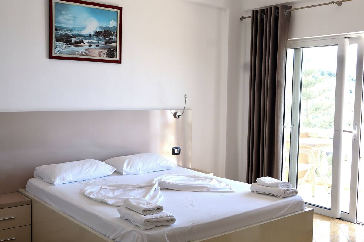 Cama bem arrumada com lençóis e toalhas brancas, um quadro na parede e vista através de uma porta de correr de vidro.
