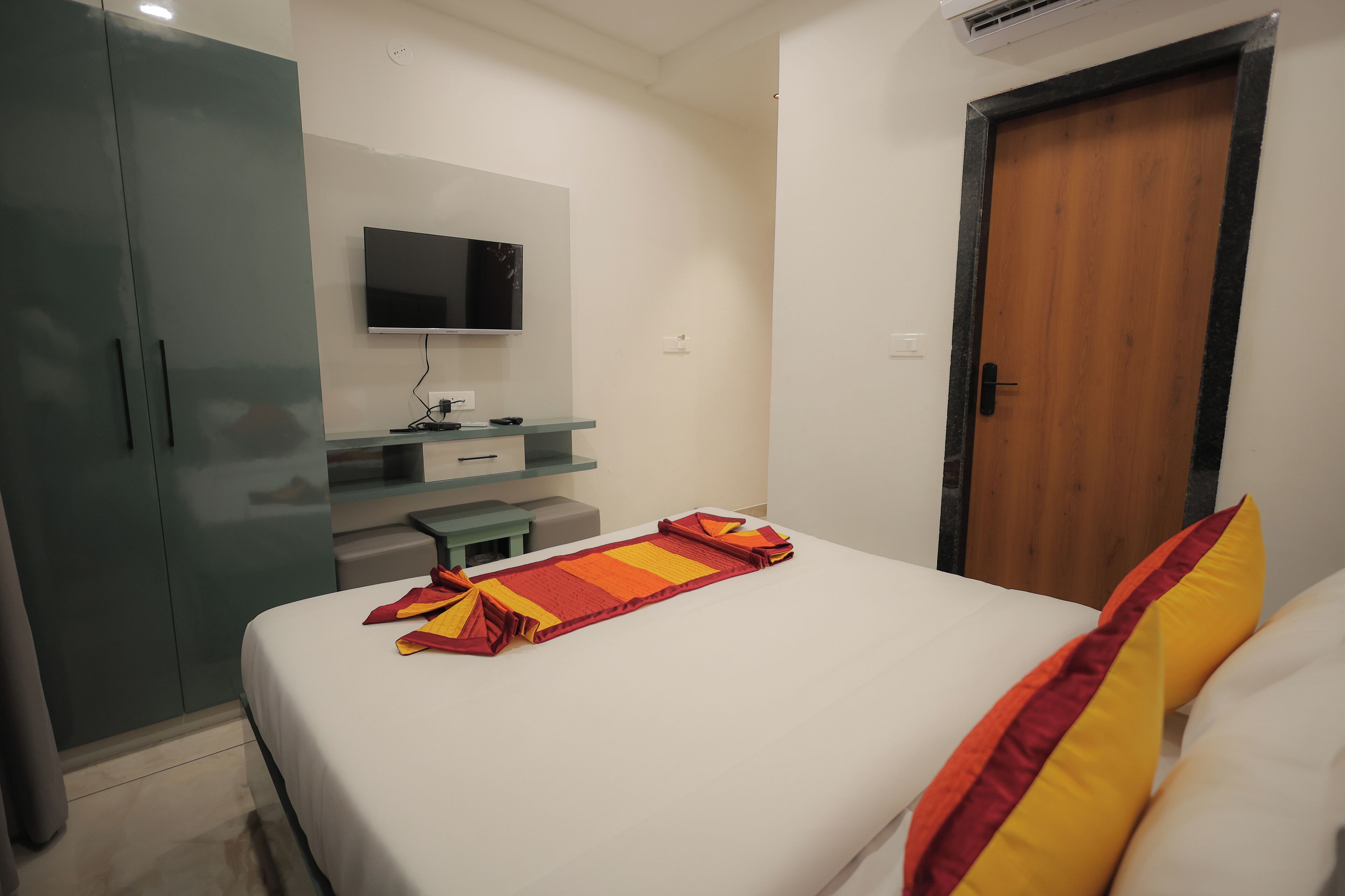 Quarto de hotel com cama, televisão na parede, porta de madeira e guarda-roupa.