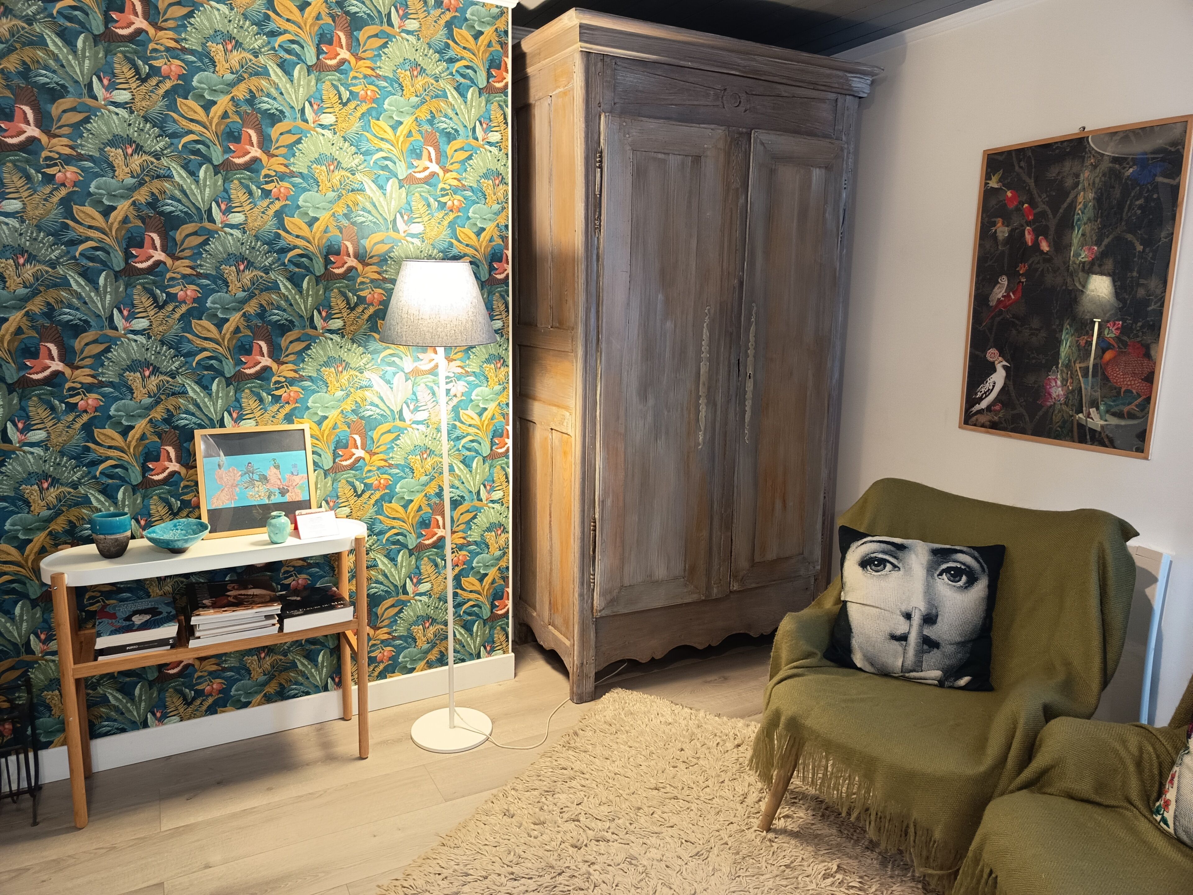 Quarto com papel de parede estampado, guarda-roupa de madeira, poltrona verde, mesa pequena com objetos decorativos e um quadro na parede.