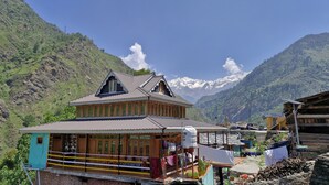 Uma casa de madeira tradicional com telhado inclinado, varanda com guarda-corpo e vista para montanhas nevadas ao fundo.