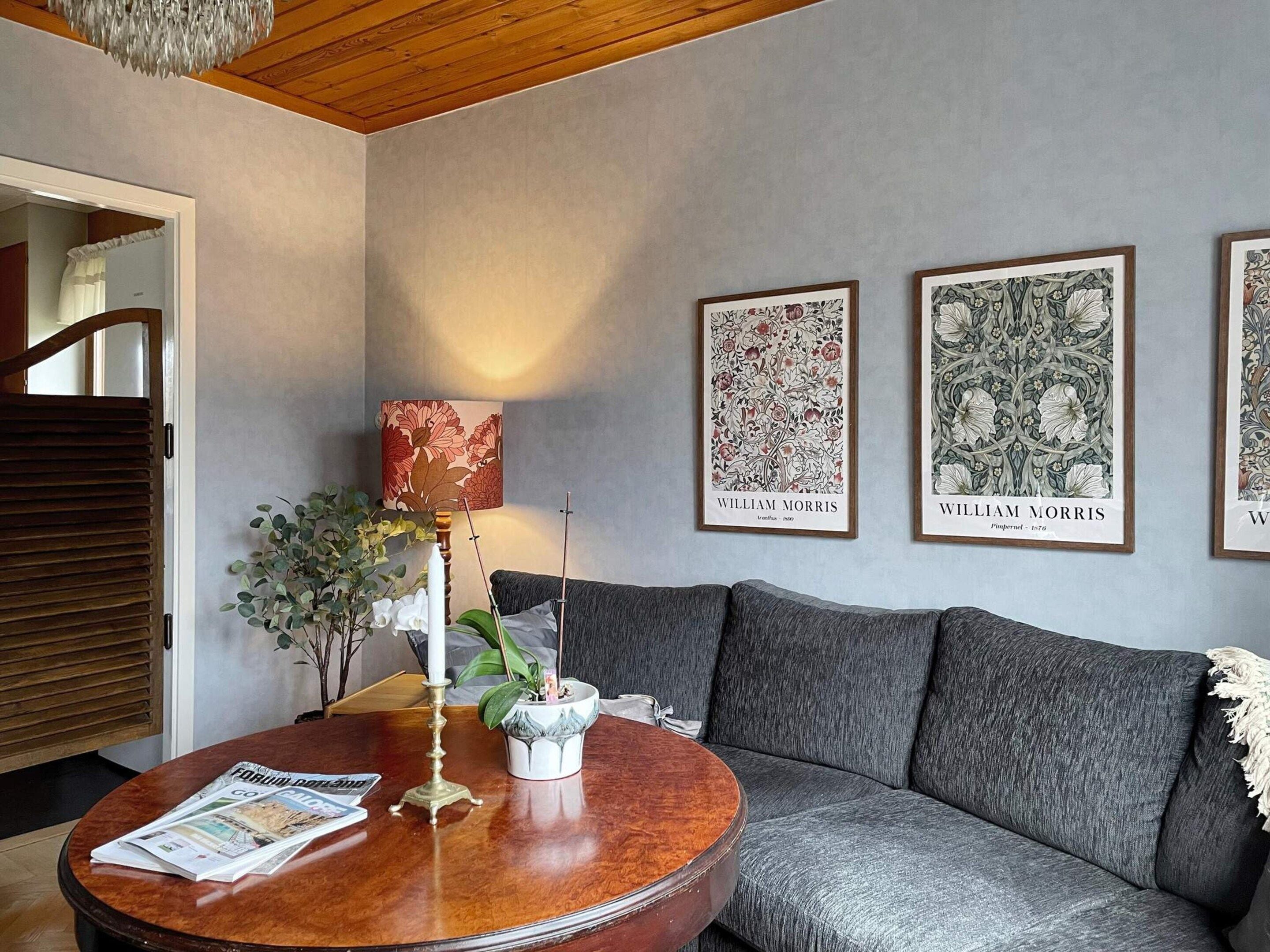 Uma sala de estar moderna com uma mesa de centro redonda de madeira, um sofá cinza e três impressões botânicas emolduradas na parede.