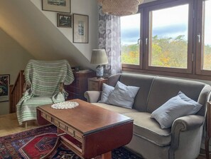 Uma sala de estar acolhedora com sofá, poltrona, mesa de madeira e janela com vista para árvores.