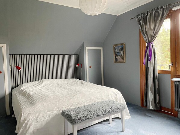 Quarto com cama, um banquinho, um quadro e janela com cortinas.