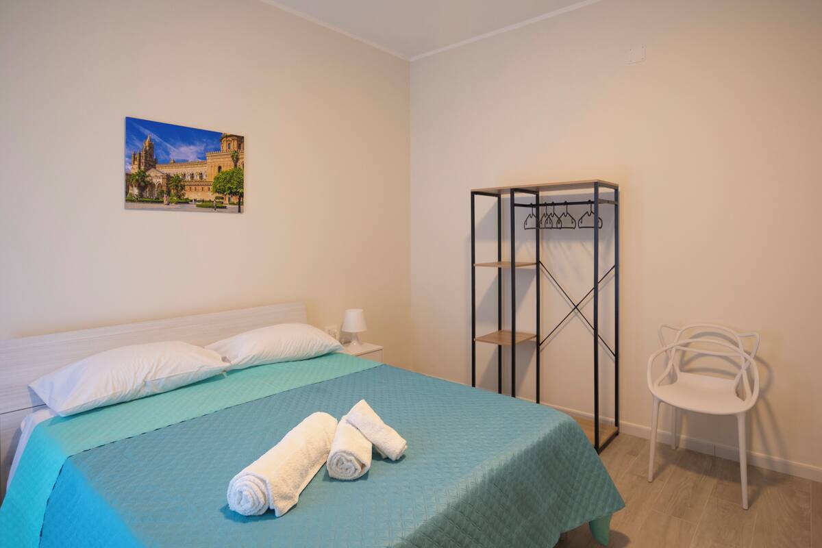 Quarto de hotel com cama, mesa de cabeceira, poltrona, cabideiro e um quadro na parede.
