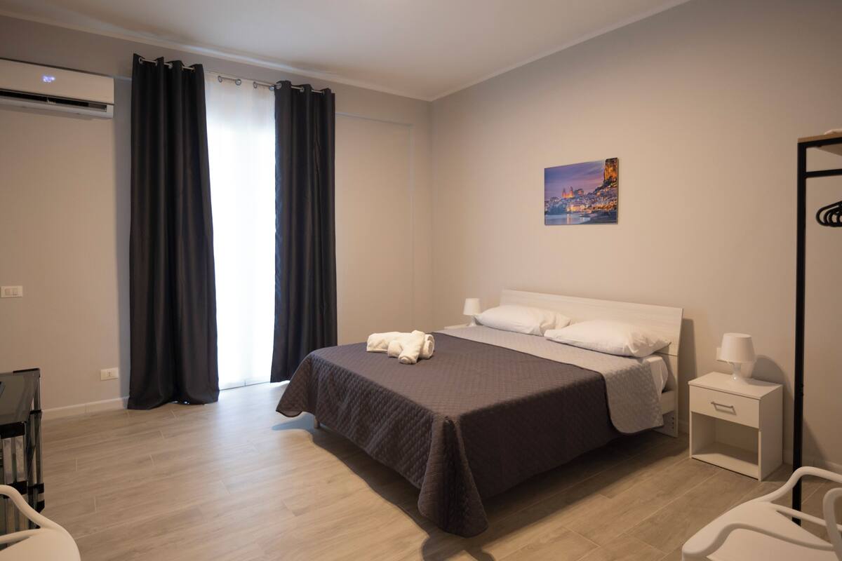 Quarto de hotel moderno com uma cama grande, uma mesa de cabeceira com abajur, uma cadeira e um quadro na parede.