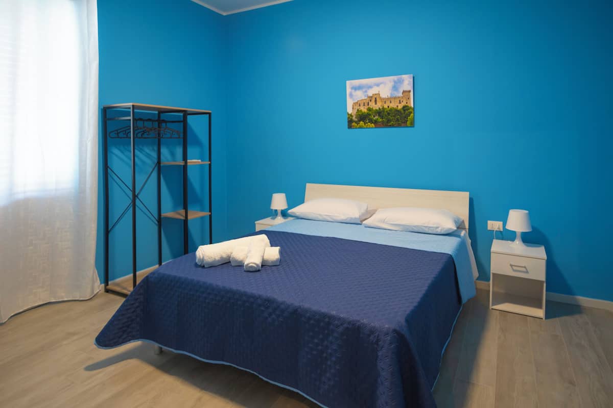 Quarto com cama, mesa de cabeceira, prateleira na parede e janela com cortina.