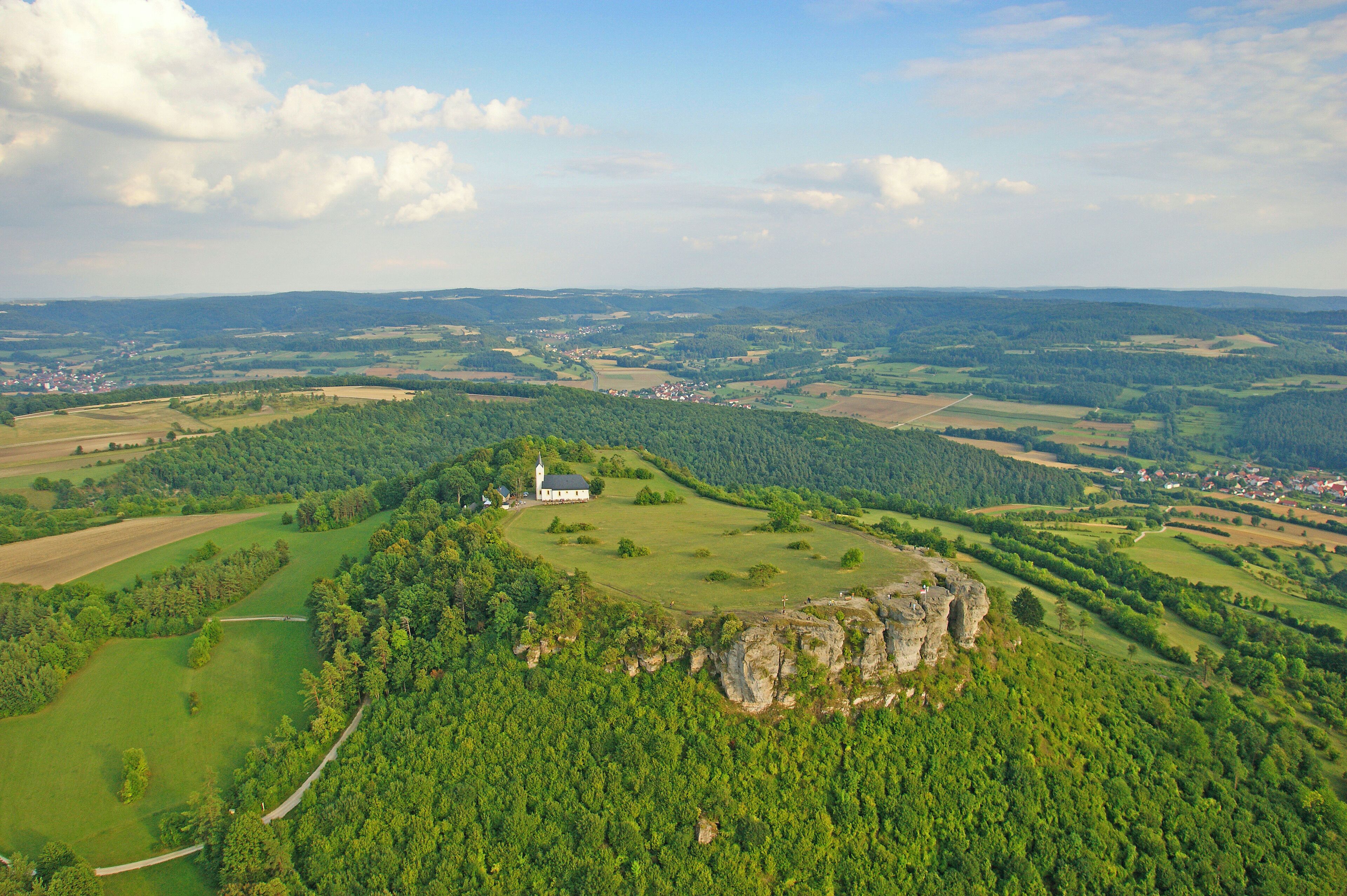 Uma vista panorâmica de uma paisagem rural com uma igreja no topo de uma colina, rodeada por campos e florestas.