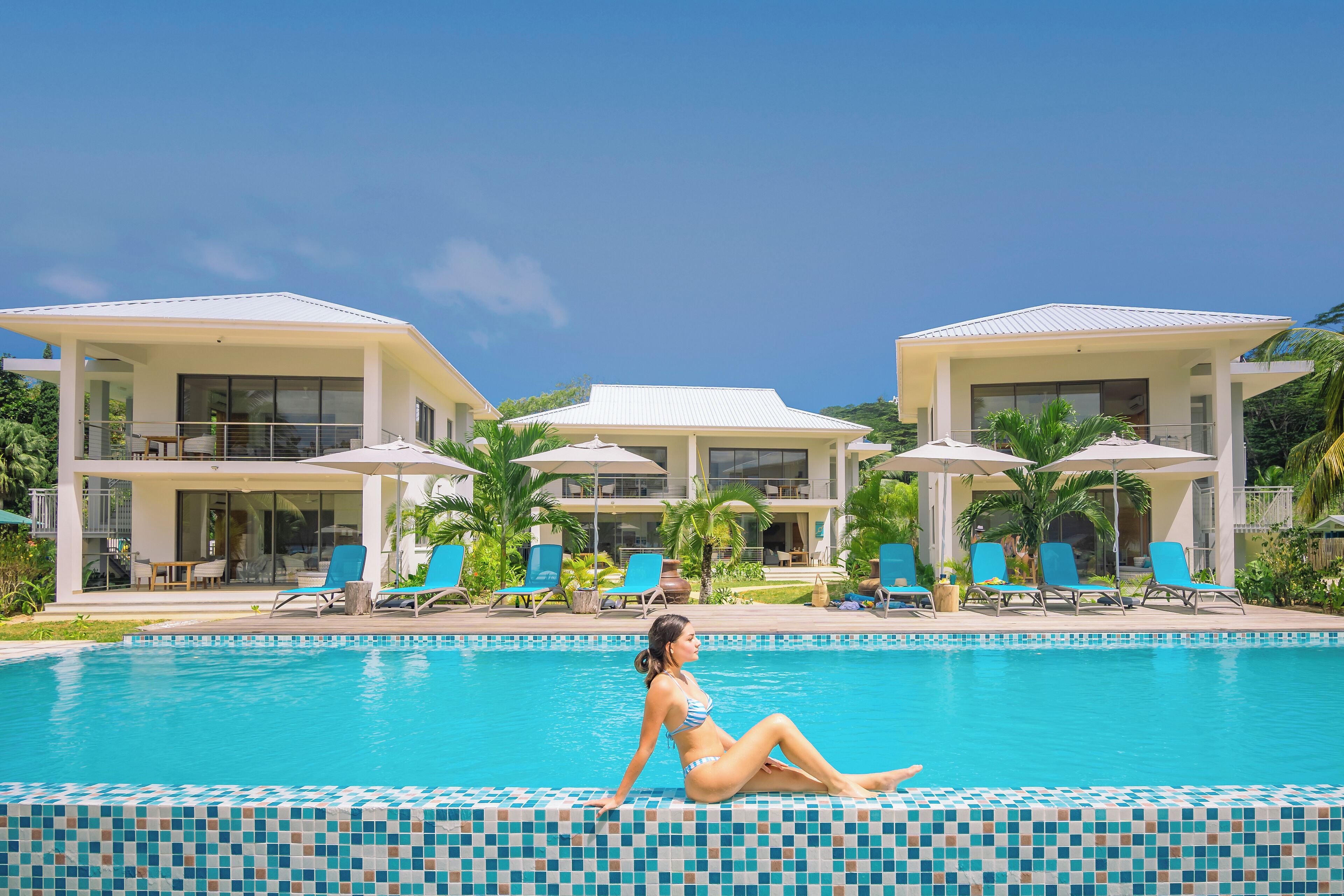 Vista da piscina com uma mulher sentada à beira da água, cadeiras de descanso, guarda-sóis e um edifício moderno com amplas janelas e borda da piscina revestida.