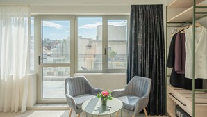 Quarto moderno com duas poltronas cinzentas, uma mesa redonda pequena com um vaso de flores, uma janela grande com vista para edifícios e um cabideiro.