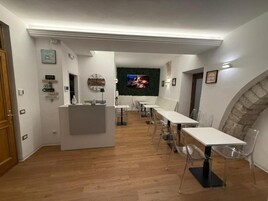 Uma área de refeições moderna com uma mesa comprida e cadeiras, um balcão de recepção pequeno e uma tela de TV exibindo uma vista urbana.