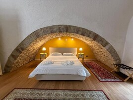Quarto com teto em arco de pedra, cama com roupa de cama branca, duas mesas de cabeceira com abajures e um tapete estampado em vermelho.