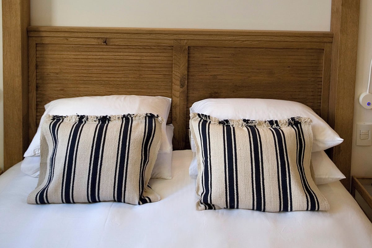 Cama com duas almofadas listradas, cabeceira de madeira e luminária fixada na parede.