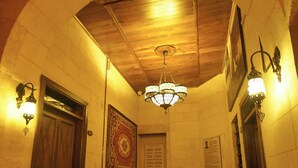 Um quarto com pouca luz, uma porta de madeira, uma mesinha, um banco estofado e um candelabro.