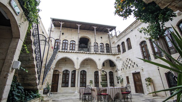 Um pátio com mesa e cadeiras, rodeado por edifícios de pedra com arcos e varandas.