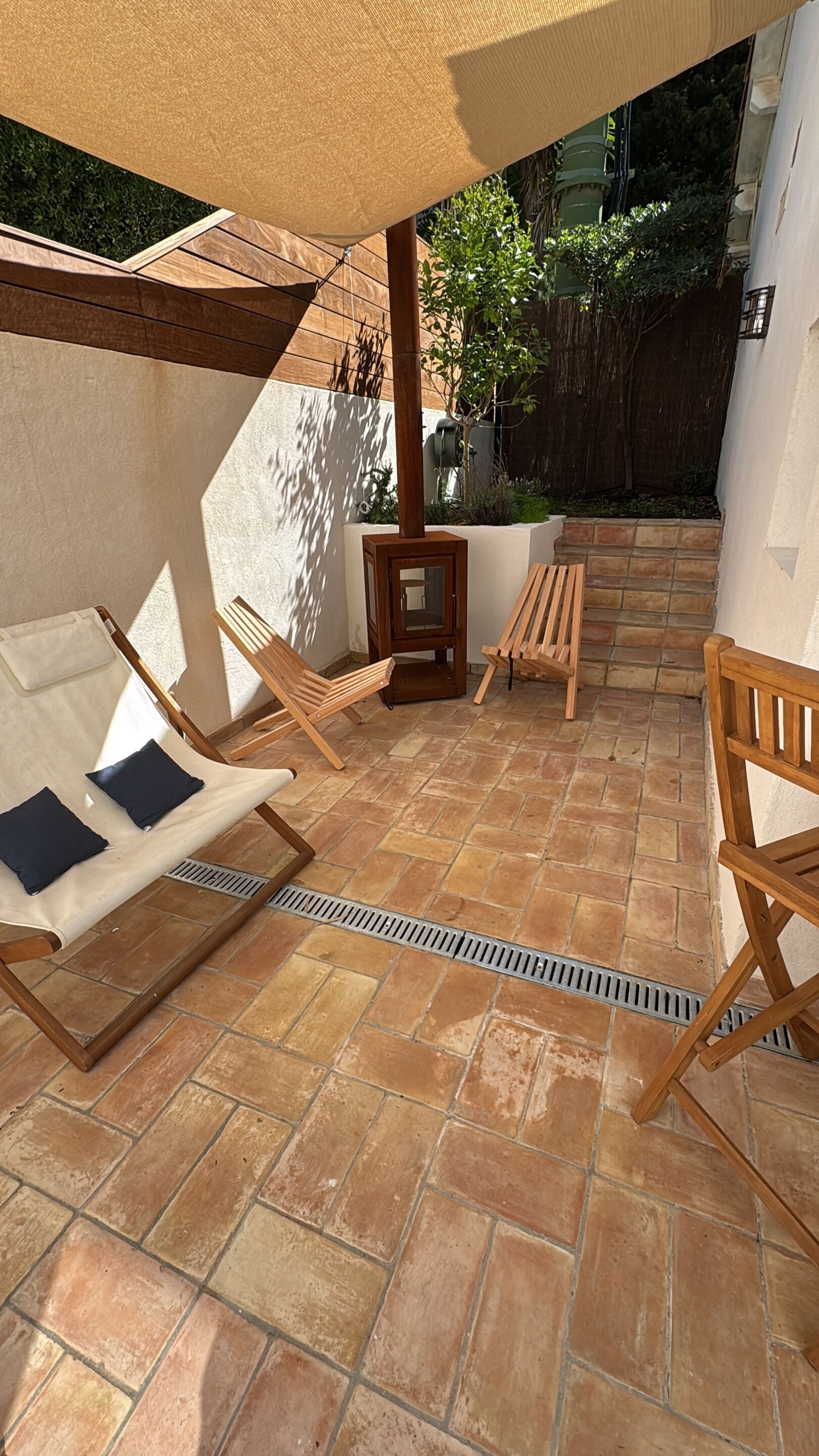 Uma área de pátio com piso de azulejo, cadeiras de madeira, uma mesa pequena e uma pérgula que oferece sombra.