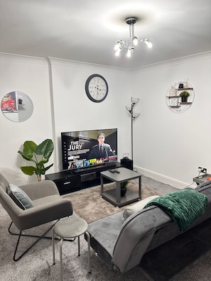 Uma sala de estar moderna com uma TV de tela plana, um sofá, uma mesa de centro, um abajur e um relógio de parede.