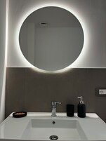 Uma casa de banho com uma pia branca, um espelho redondo com luz e um dispensador de sabonete preto.