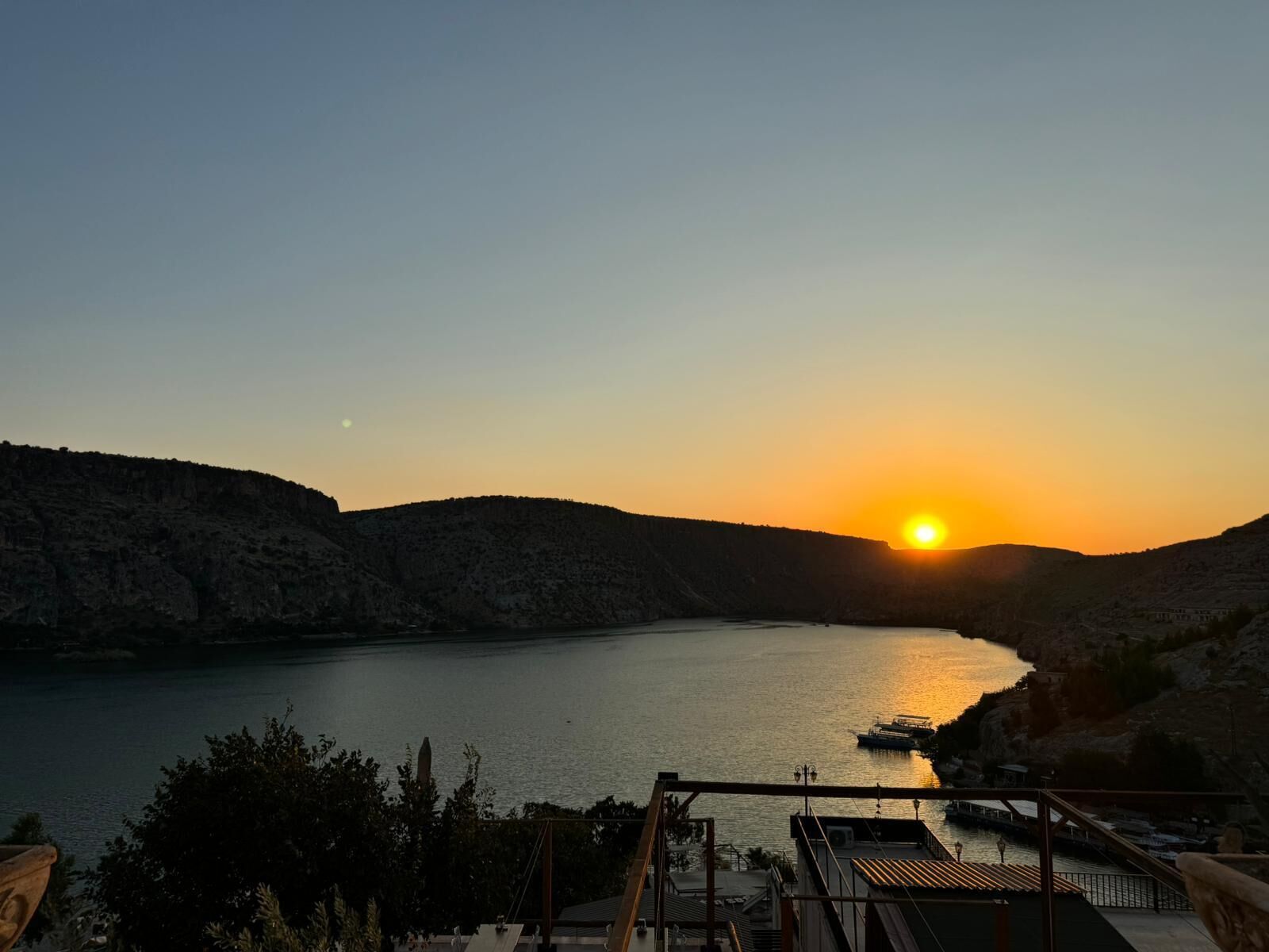 Um pôr do sol sobre um lago calmo, com um barco atracado à direita, cercado por colinas rochosas.
