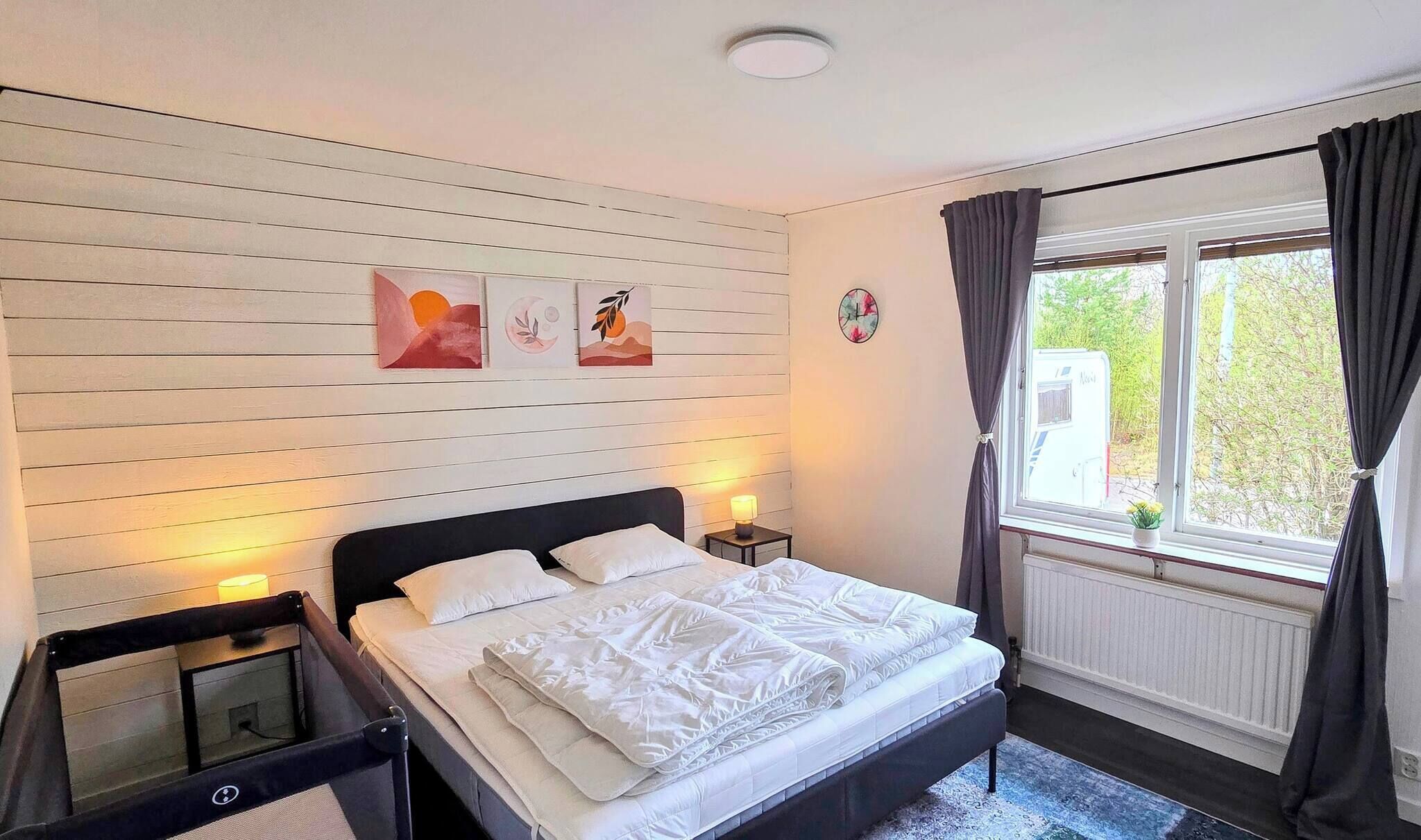 Quarto com cama, mesa de cabeceira, janela com cortinas e quadros nas paredes.