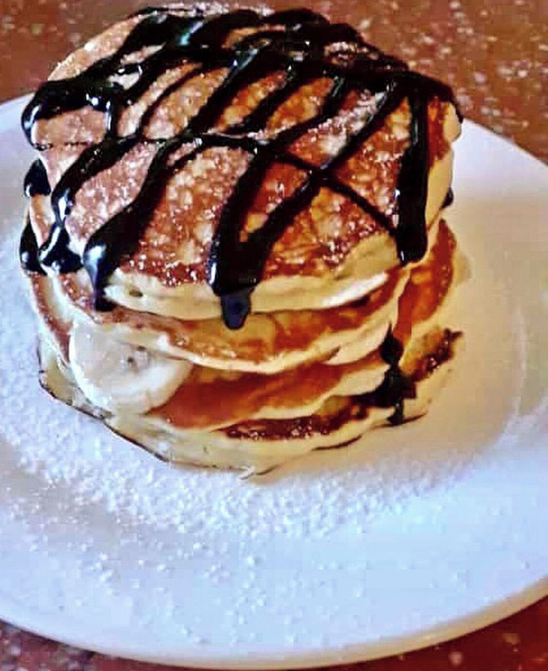 Uma pilha de panquecas com calda de chocolate e açúcar em pó.