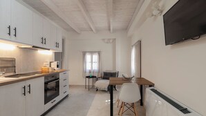 Cozinha moderna com armários brancos, bancada de madeira e forno embutido. Há uma área de refeições com mesa de madeira e cadeiras brancas. Uma televisão está fixada na parede, e um radiador é visível no lado direito.